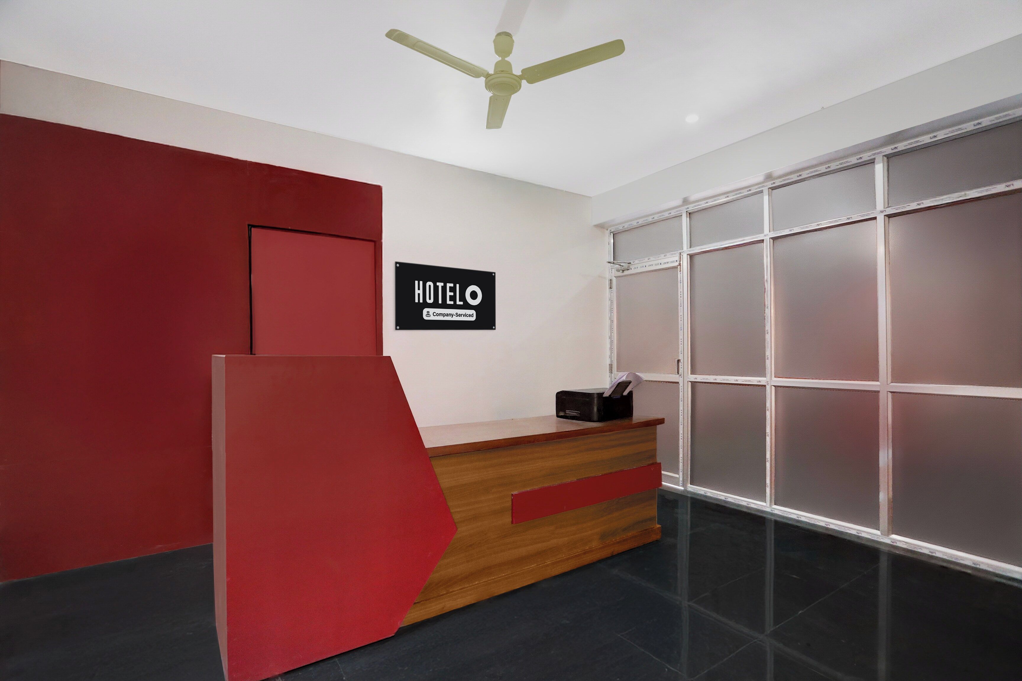 Uma área de recepção de hotel com um balcão vermelho, uma placa indicando serviço empresarial, um ventilador de teto e uma grande porta de vidro fosco.