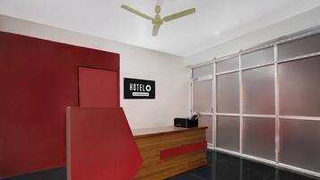 Uma área de recepção de hotel com um balcão vermelho, uma placa indicando serviço empresarial, um ventilador de teto e uma grande porta de vidro fosco.
