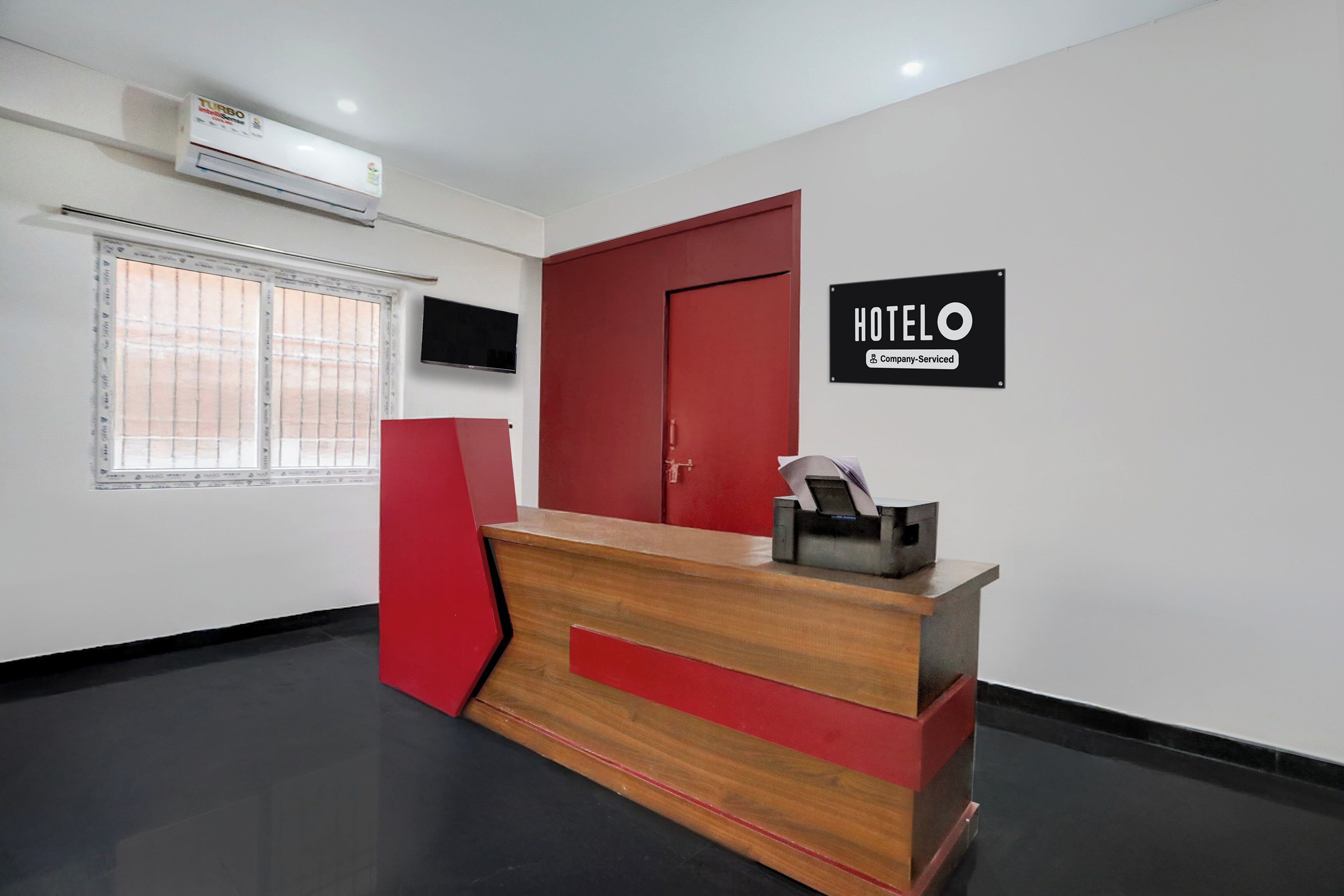 Balcão de recepção de hotel com esquema de cores vermelho e preto, uma televisão de tela plana na parede e uma placa indicando que o hotel é para uso corporativo.