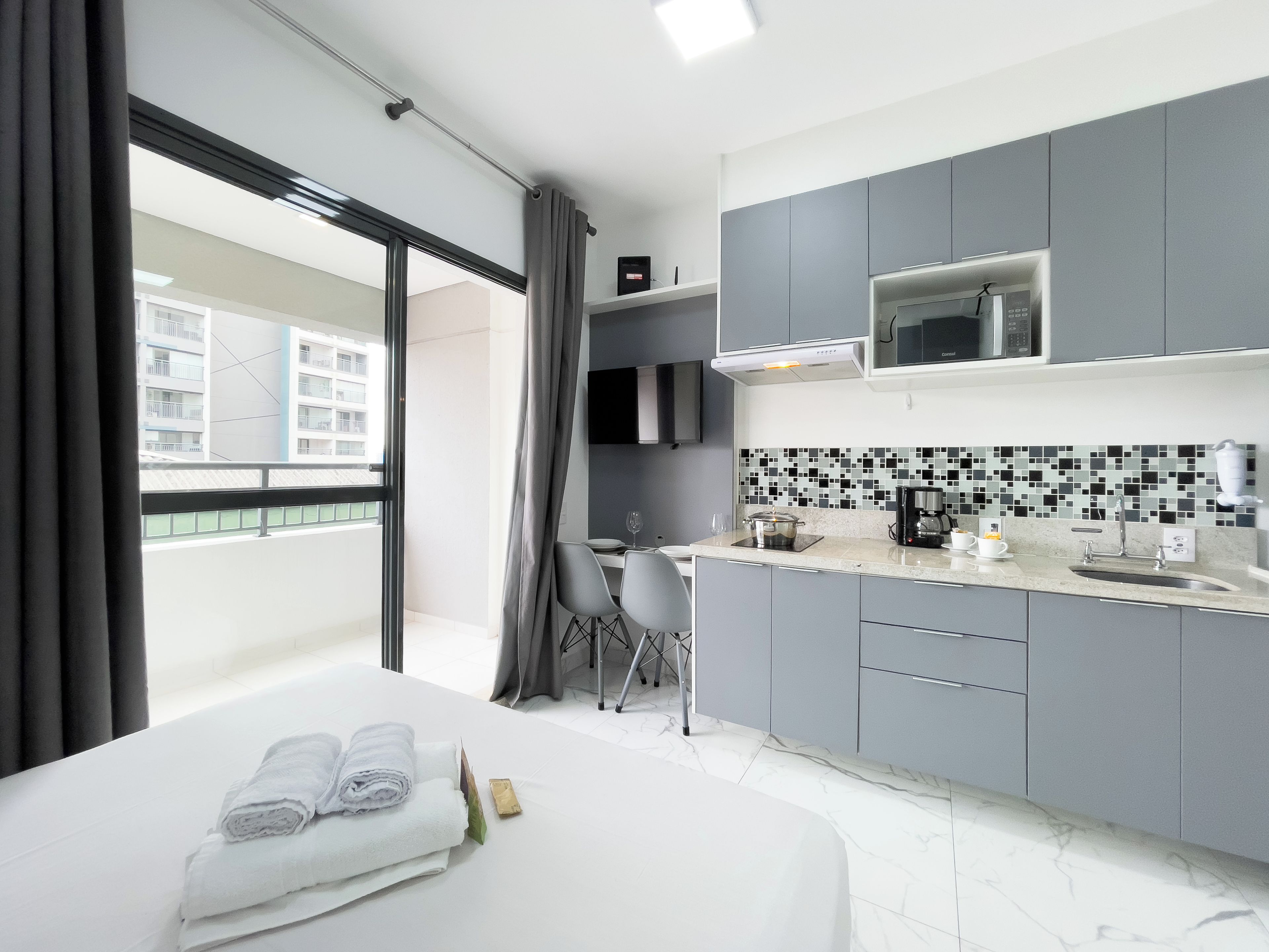 Quarto de hotel moderno com varanda, cama com toalhas, cozinha compacta e televisão de tela plana.