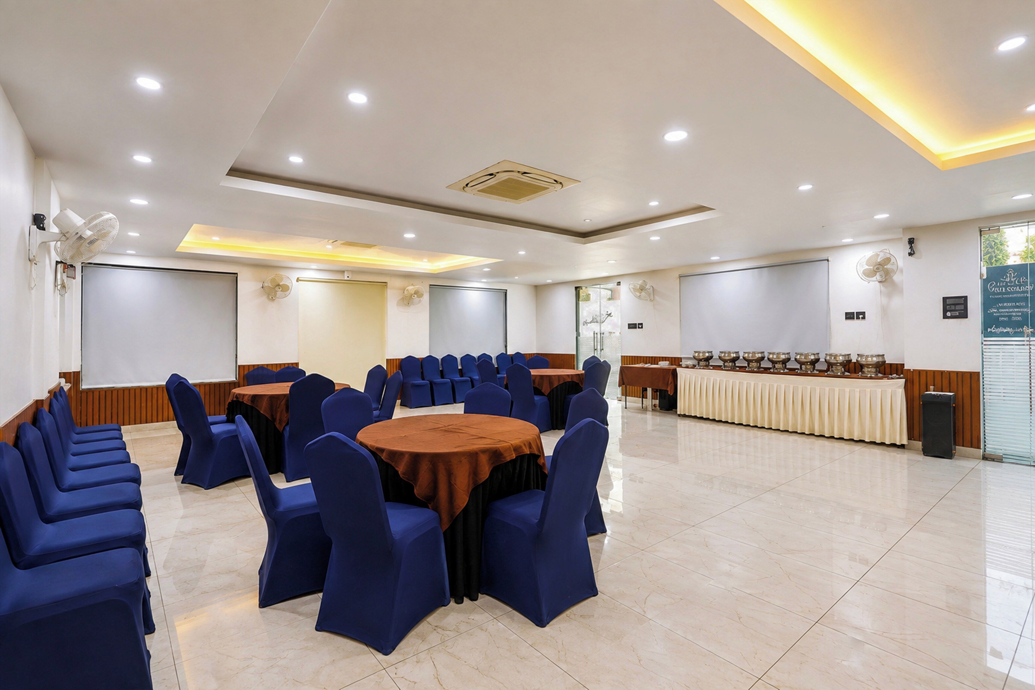 Sala de conferências com mesas redondas e cadeiras azuis, equipada com ventiladores de teto e iluminação embutida.