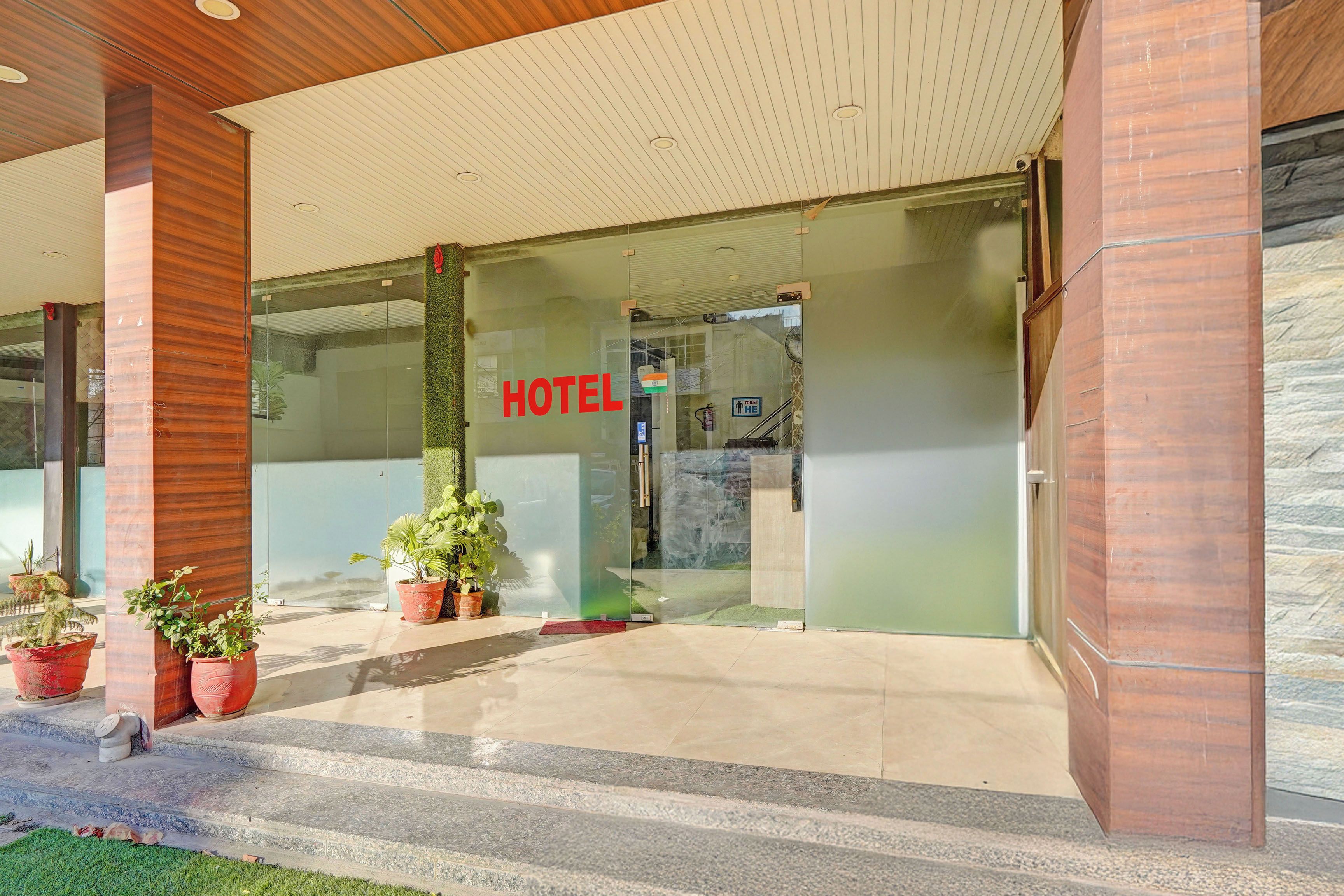 Entrada de hotel com porta de vidro, plantas em vasos e placa indicando um banheiro.