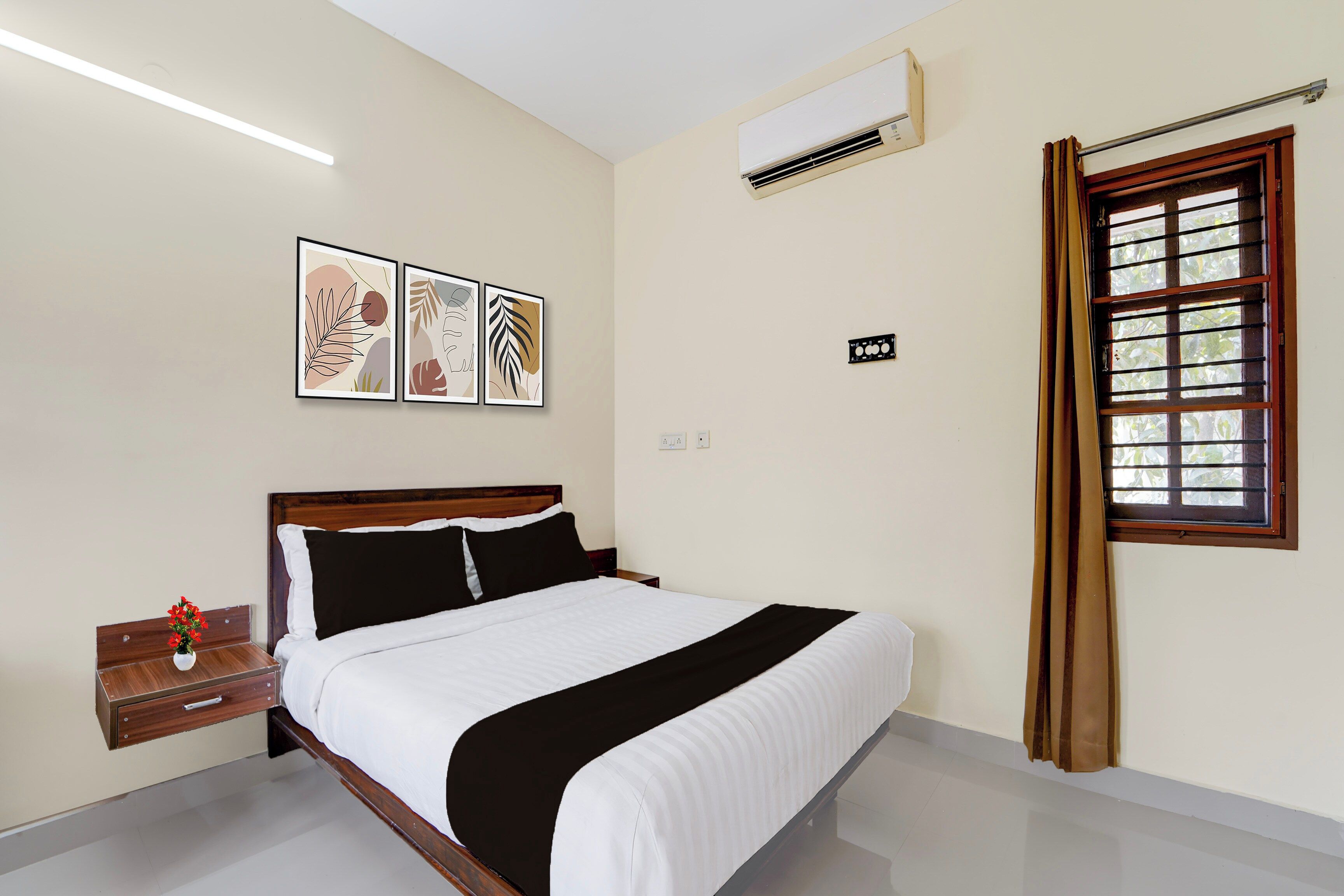 Quarto de hotel com cama, mesa de cabeceira, vaso com flores, janela com cortinas e ar-condicionado fixado na parede.