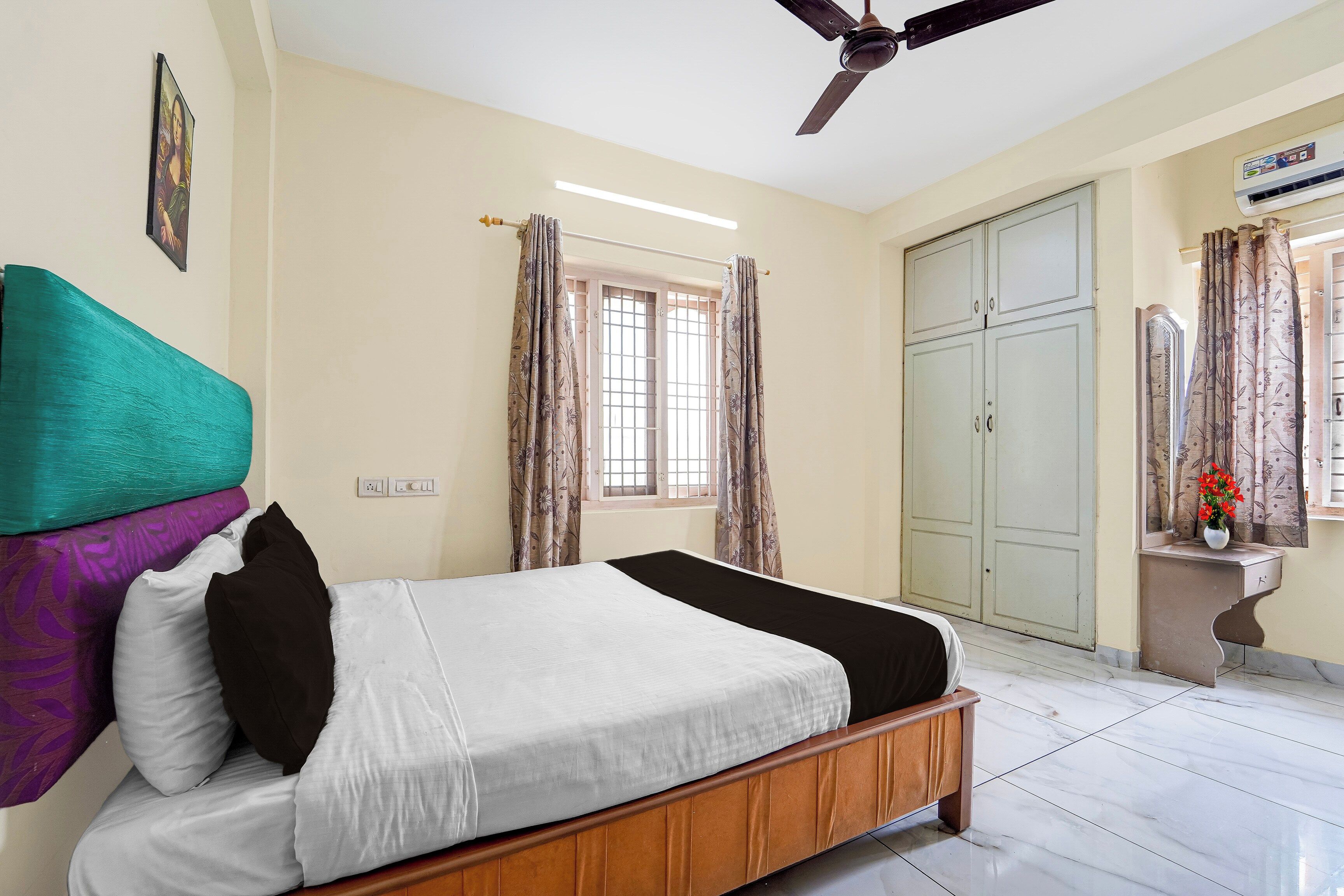 Quarto com cama, ventoinha no teto, janela com cortinas, guarda-roupa e aparelho de ar condicionado.