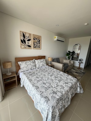 Quarto com cama grande, mesa de cabeceira com abajur, sofá, espelho e obras de arte nas paredes.
