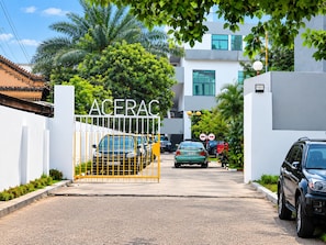 Uma entrada fechada por portão com uma placa indicando 'ACERAC', que leva a um edifício moderno com fachada branca e várias janelas.