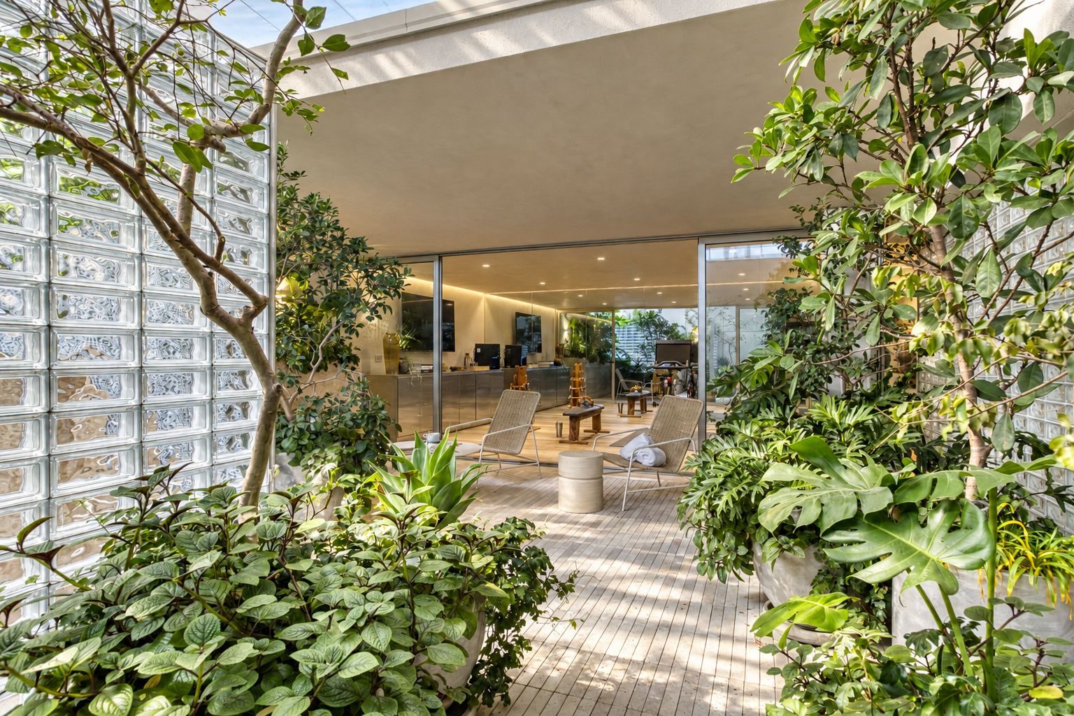 Espaço interior com design moderno, apresentando área de estar com cadeiras e mesa de jantar, cercada por plantas verdes exuberantes e uma parede de vidro com desenhos decorativos.