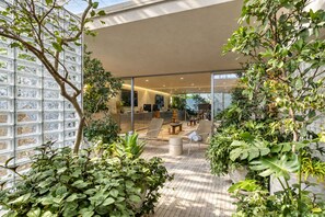 Espaço interior com design moderno, apresentando área de estar com cadeiras e mesa de jantar, cercada por plantas verdes exuberantes e uma parede de vidro com desenhos decorativos.