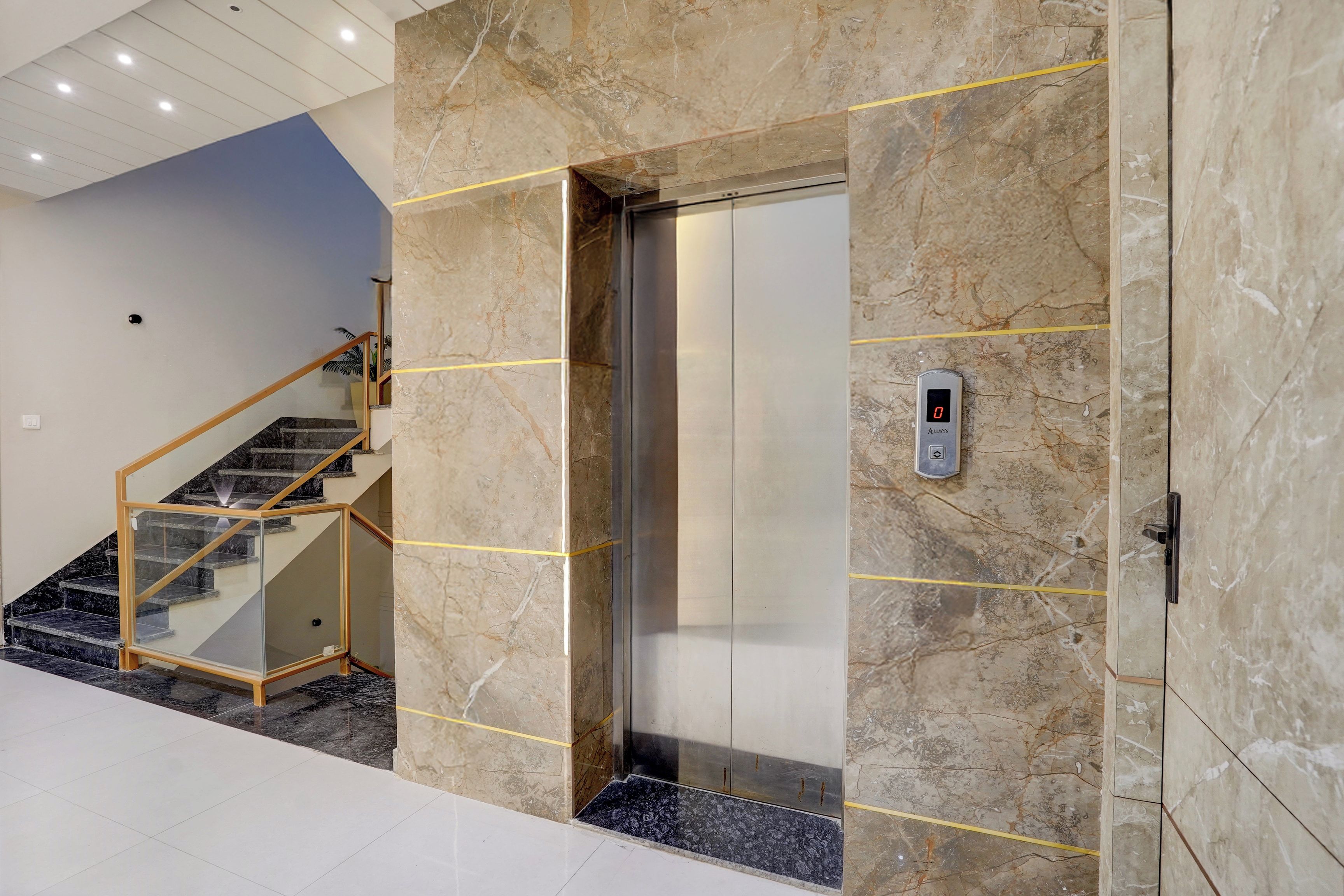 Um elevador com visor digital, parede de mármore e escada com guarda-corpo de vidro.