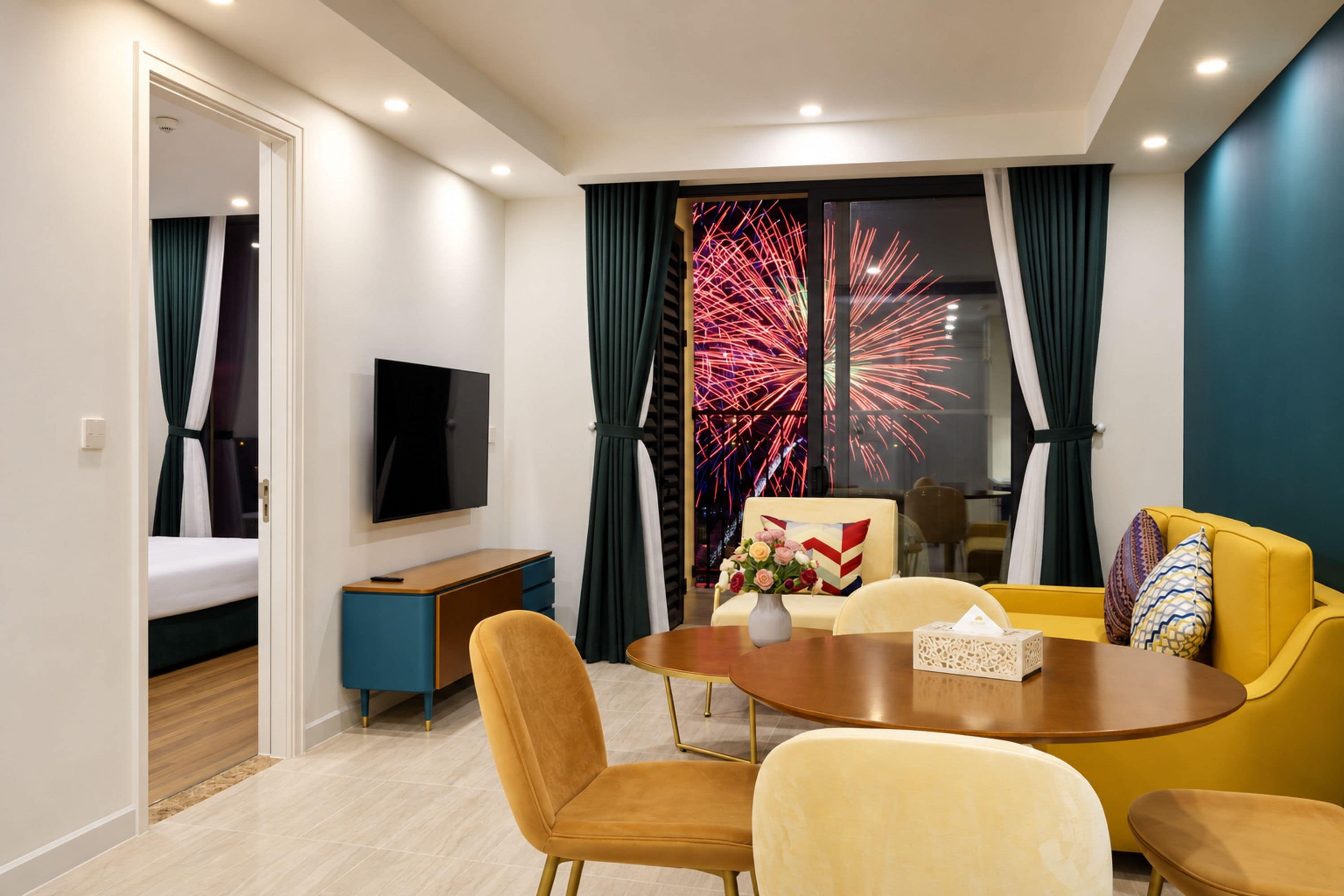 Quarto de hotel moderno com cama, televisão, área de refeições com mesa e cadeiras, e uma janela grande com cortinas.