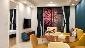 Quarto de hotel moderno com cama, televisão, área de refeições com mesa e cadeiras, e uma janela grande com cortinas.