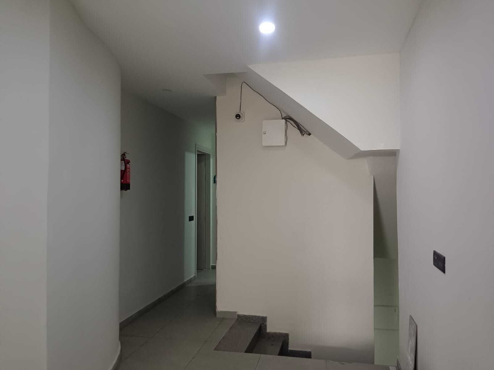 Corredor com escada que leva a um andar superior, detector de incêndio na parede e uma porta no final.