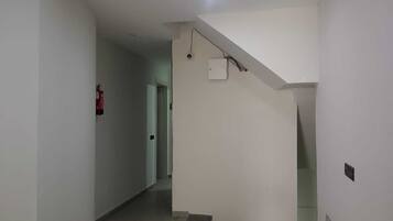 Corredor com escada que leva a um andar superior, detector de incêndio na parede e uma porta no final.