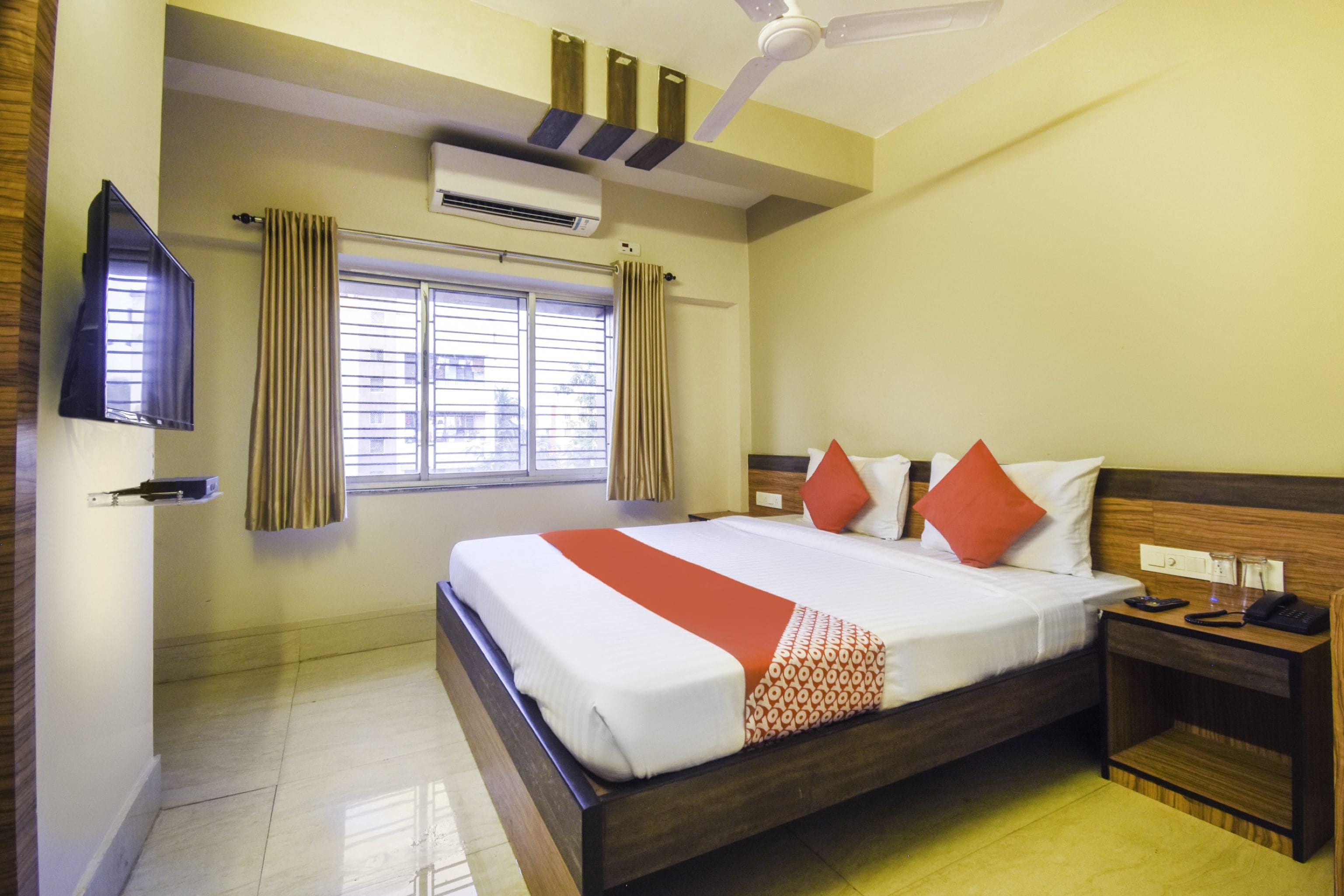 Quarto de hotel com cama, televisão, ar condicionado, janela com cortinas e ventilador de teto.
