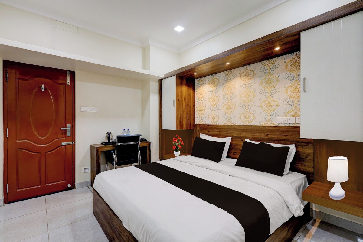 Quarto de hotel com cama grande, cabeceira de madeira, escrivaninha com cadeira, mesa de cabeceira com abajur e porta vermelha.
