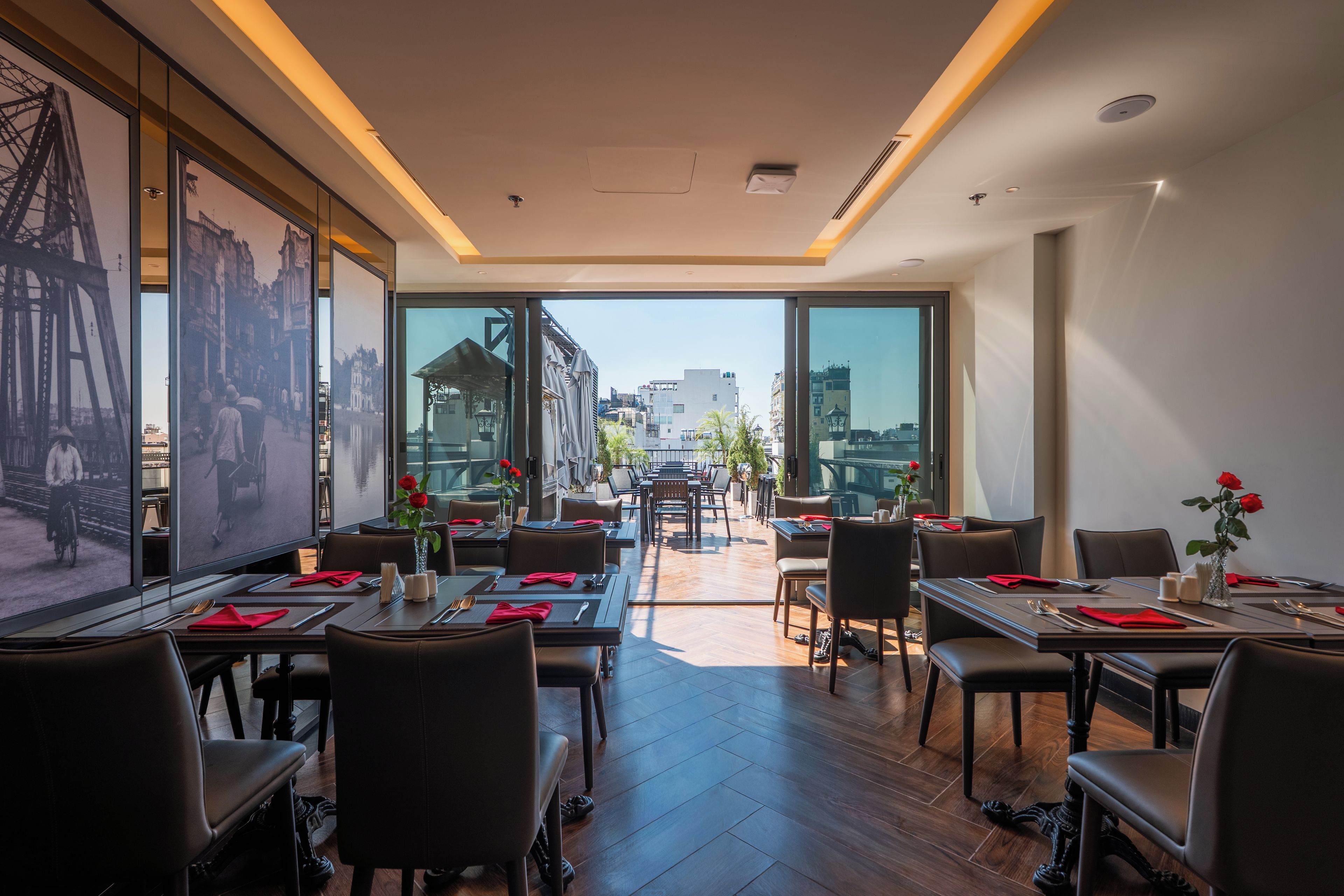 Um restaurante com mesas preparadas para refeições, grandes janelas com vista para a cidade e obras de arte nas paredes.