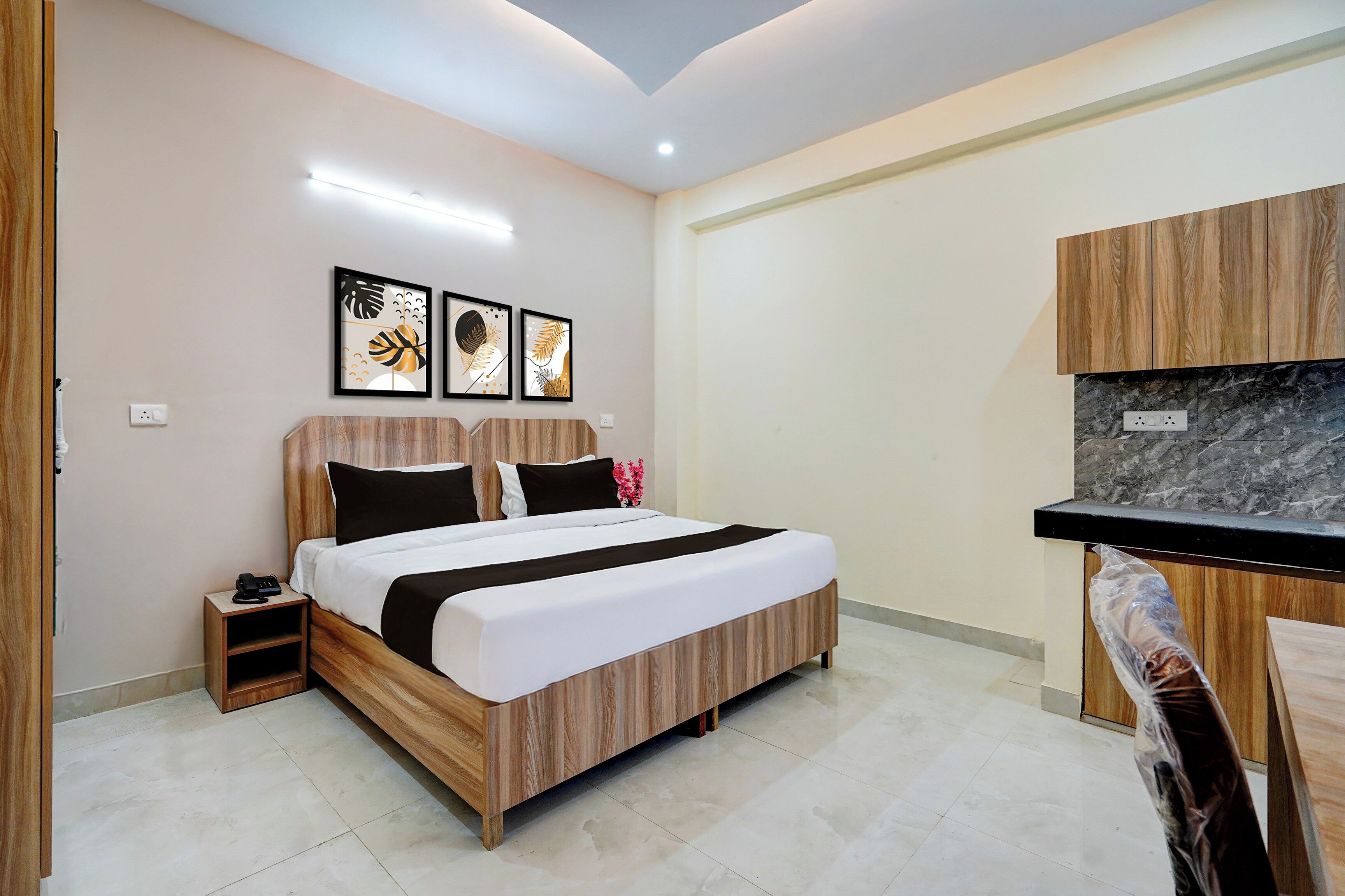 Quarto de hotel com cama, mesa de cabeceira e pequena escrivaninha com cadeira. Quadros emoldurados decoram a parede e um armário de madeira possui bancada de mármore.