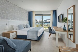 Quarto de hotel moderno com cama, escrivaninha, cadeira, televisão e vista para uma piscina e edifícios.
