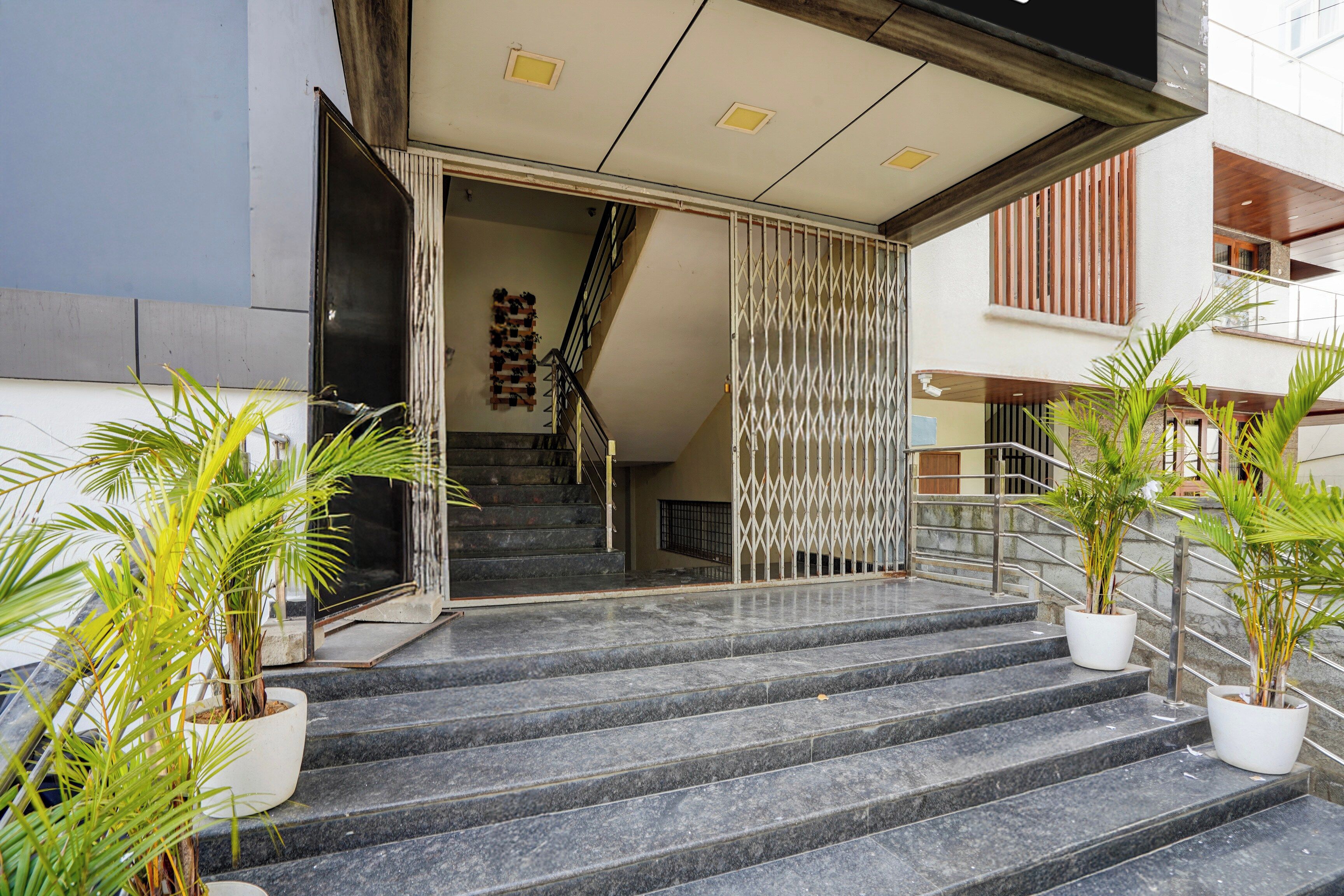 Escadaria que leva à entrada de um edifício, com portão de metal, plantas em vasos e outra escadaria de madeira ao fundo.
