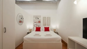 Quarto de hotel com cama, mesas de cabeceira, televisão, espelho e prateleira com quadros.