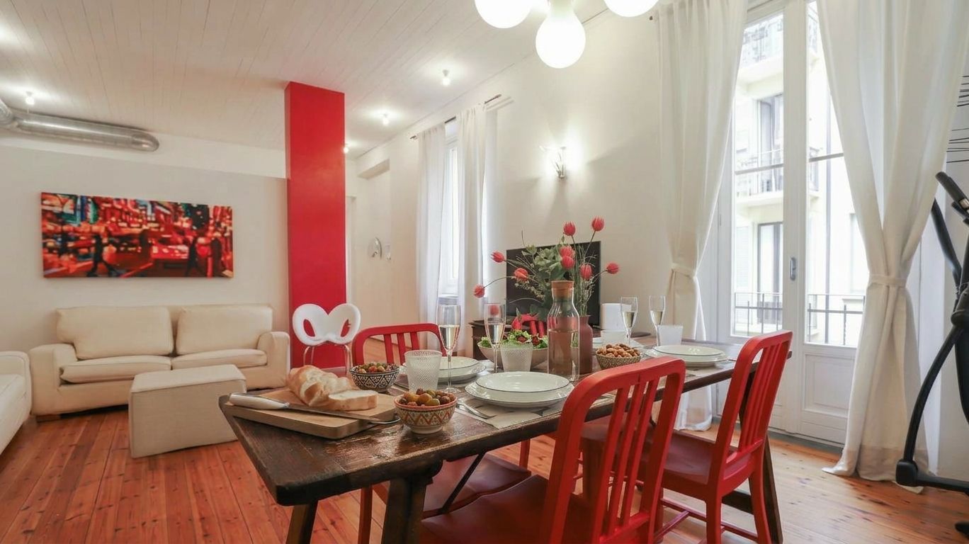 Uma sala de jantar com mesa de madeira posta para quatro pessoas, cadeiras vermelhas e um sofá branco ao fundo.