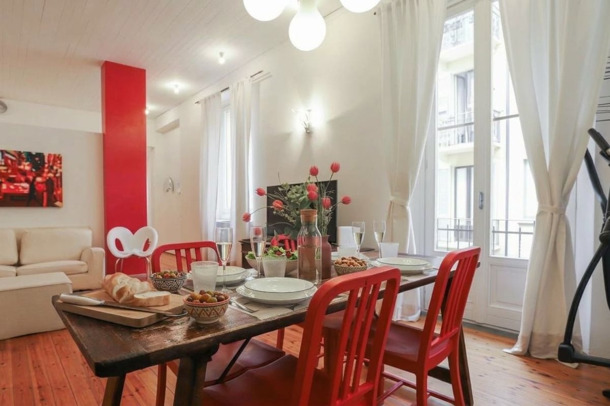 Uma sala de jantar com mesa de madeira posta para quatro pessoas, cadeiras vermelhas e um sofá branco ao fundo.