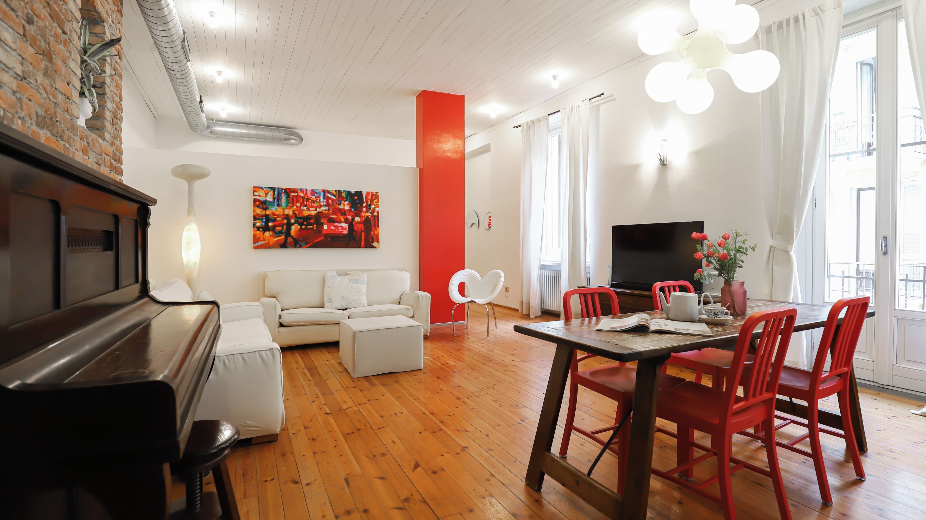 Uma sala de estar com um piano, um sofá, uma mesa de jantar com cadeiras vermelhas, uma televisão e um quadro na parede.