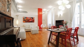 Uma sala de estar com um piano, um sofá, uma mesa de jantar com cadeiras vermelhas, uma televisão e um quadro na parede.
