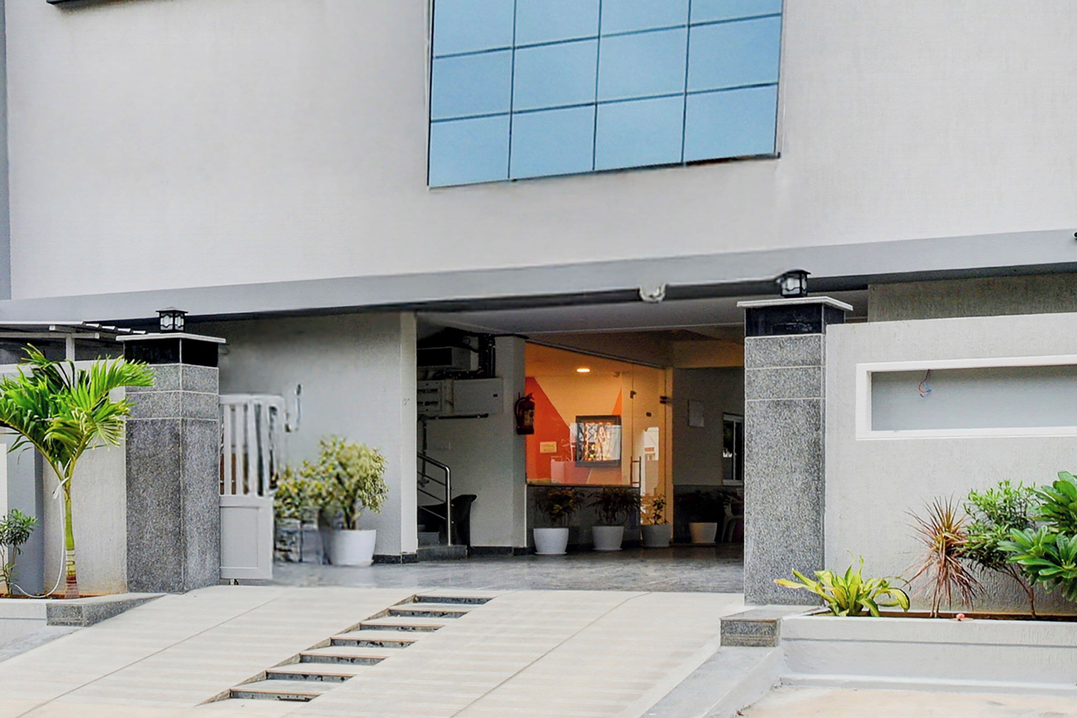 Entrada de um edifício moderno com caminho de azulejos que leva a uma porta, plantas em vasos e interior visível com uma placa vermelha.