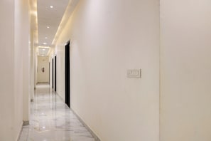 Um corredor comprido e estreito com paredes brancas e chão de mármore. Há portas de um lado e um extintor de incêndio fixado na parede.