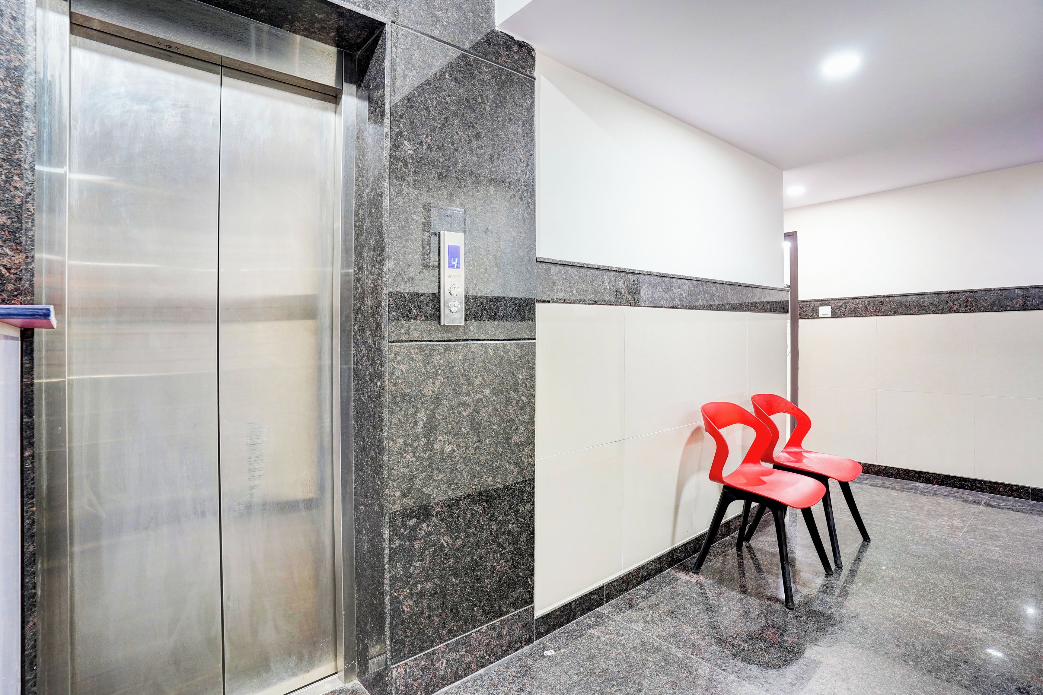 Elevador moderno com painel digital, parede de mármore e área de estar com cadeiras vermelhas.