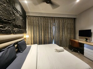 Quarto de hotel com cama, escrivaninha, televisão e cortina.