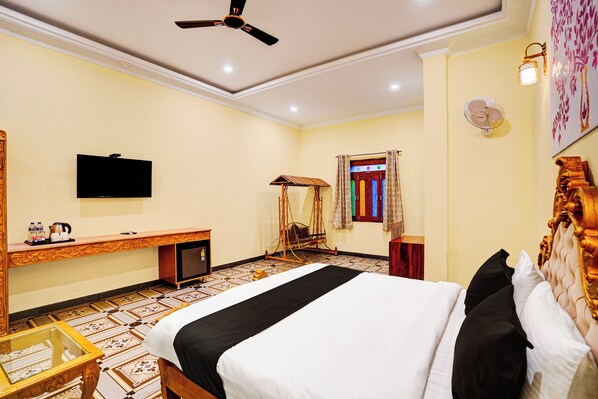 Quarto de hotel com cama, escrivaninha, televisão, ventoinha no teto e janela com cortinas.