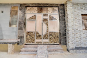 Entrada de um edifício com um portão de metal decorativo, degraus que conduzem a ele e uma parede com uma janela à esquerda.