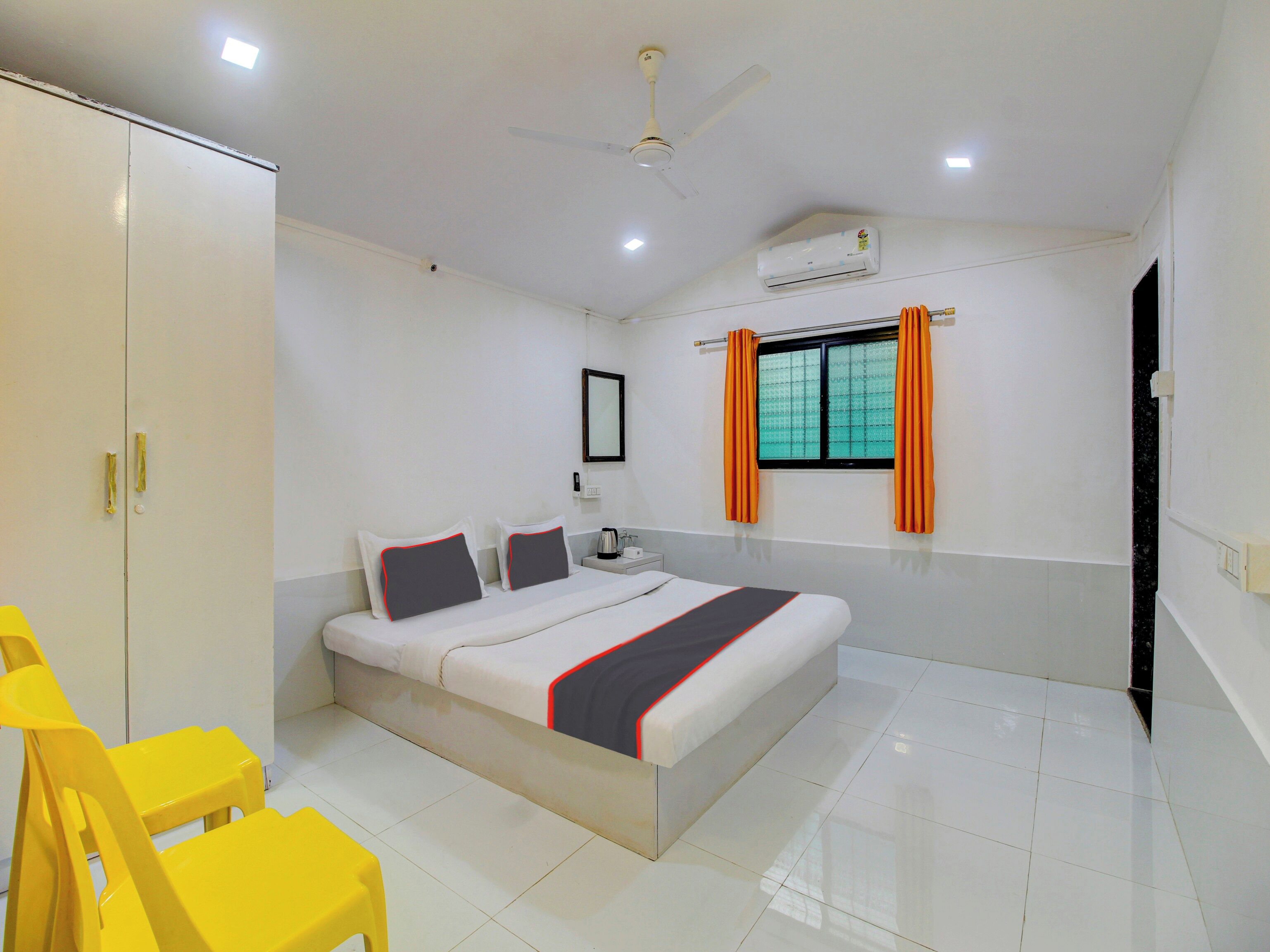 Quarto de hotel com cama, guarda-roupa, ventoinha no teto, ar condicionado, janela com cortinas e pequena mesa com luminária.