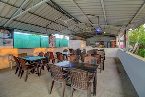 Uma área de refeições coberta ao ar livre, com mesas e cadeiras, ventilador de teto e vista para o exterior.