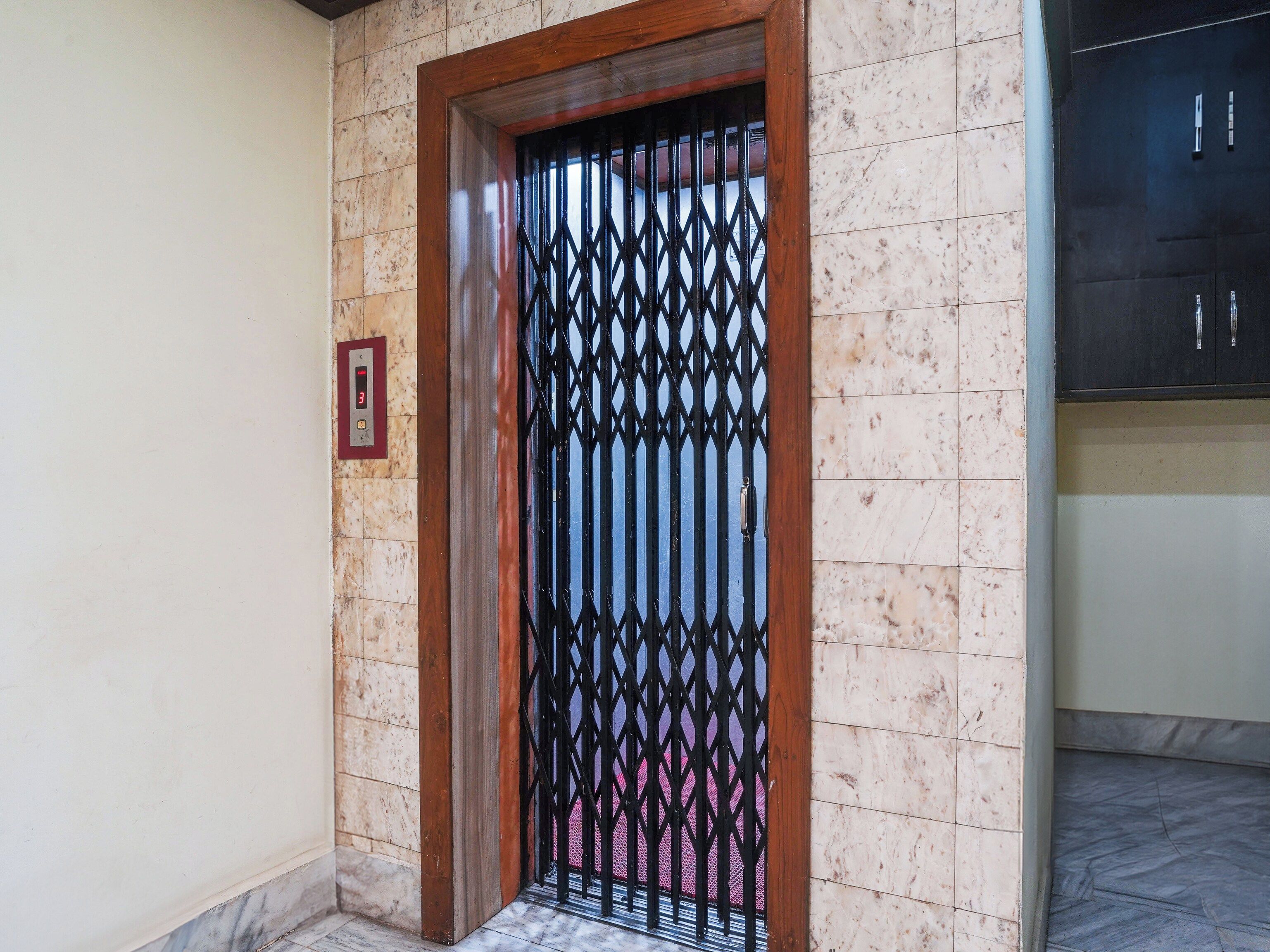 Uma porta com portão de segurança metálico, painel de controle de elevador fixado na parede e piso revestido com azulejos.