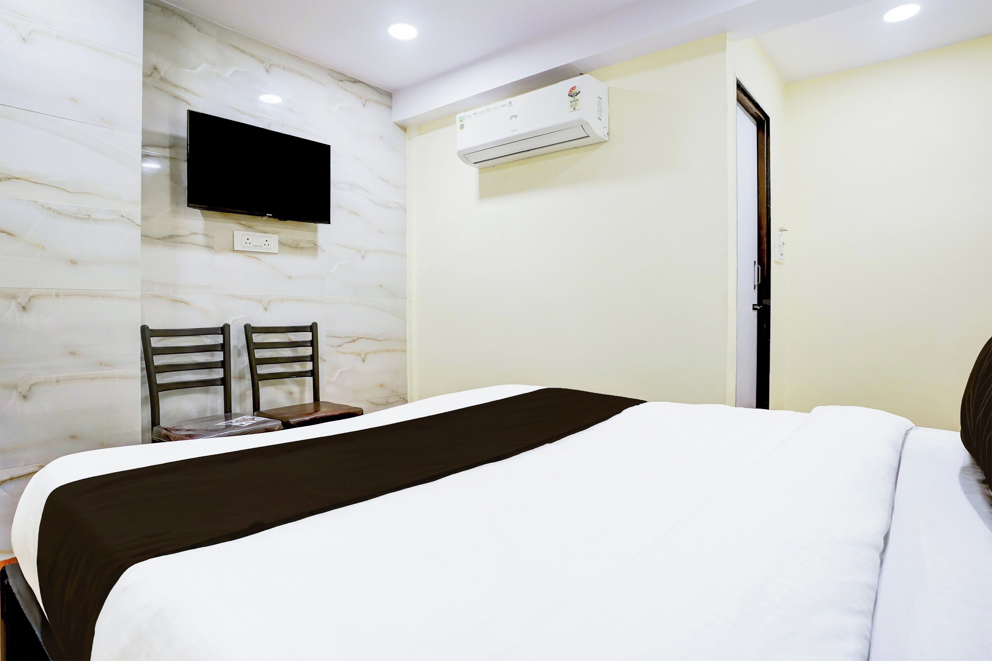 Quarto de hotel com cama, televisão de tela plana na parede, ar-condicionado, duas cadeiras e uma porta.