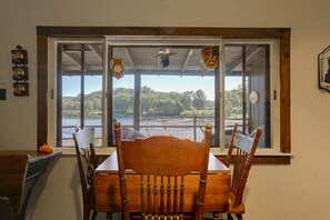 Uma área de jantar com mesa e cadeiras, uma janela com vista para um lago e árvores, e teto de madeira.