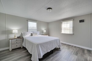 Quarto com cama, duas mesas de cabeceira, um criado-mudo, luminária na parede e duas janelas com persianas.