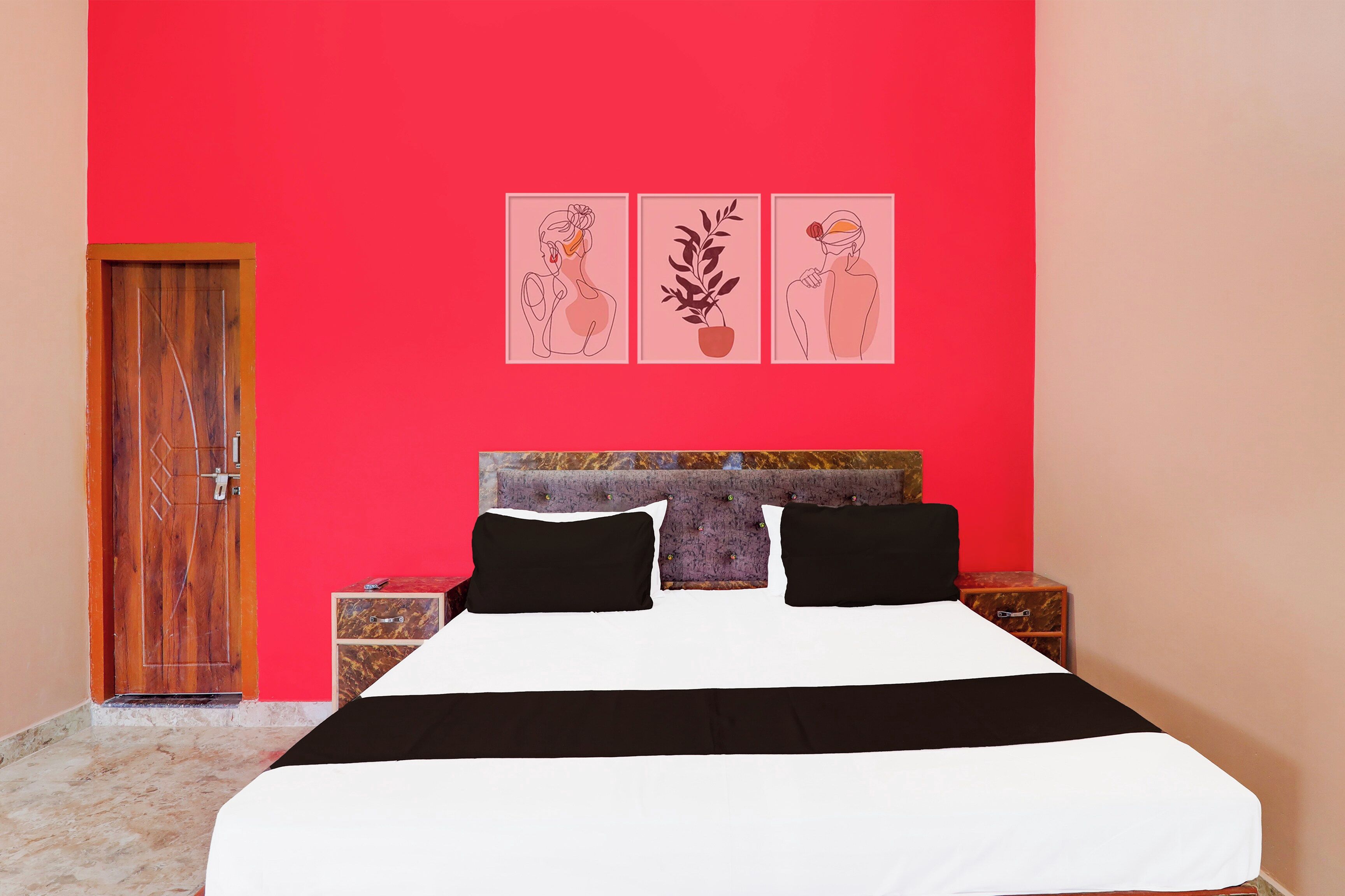 Quarto com parede vermelha, porta de madeira, cama com roupa de cama preta e branca e três quadros acima da cama.