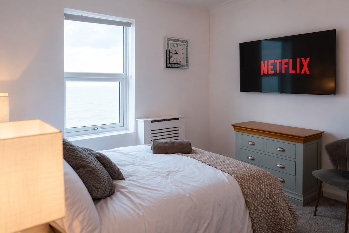 Quarto com cama, cômoda, cadeira, televisão na parede e janela.