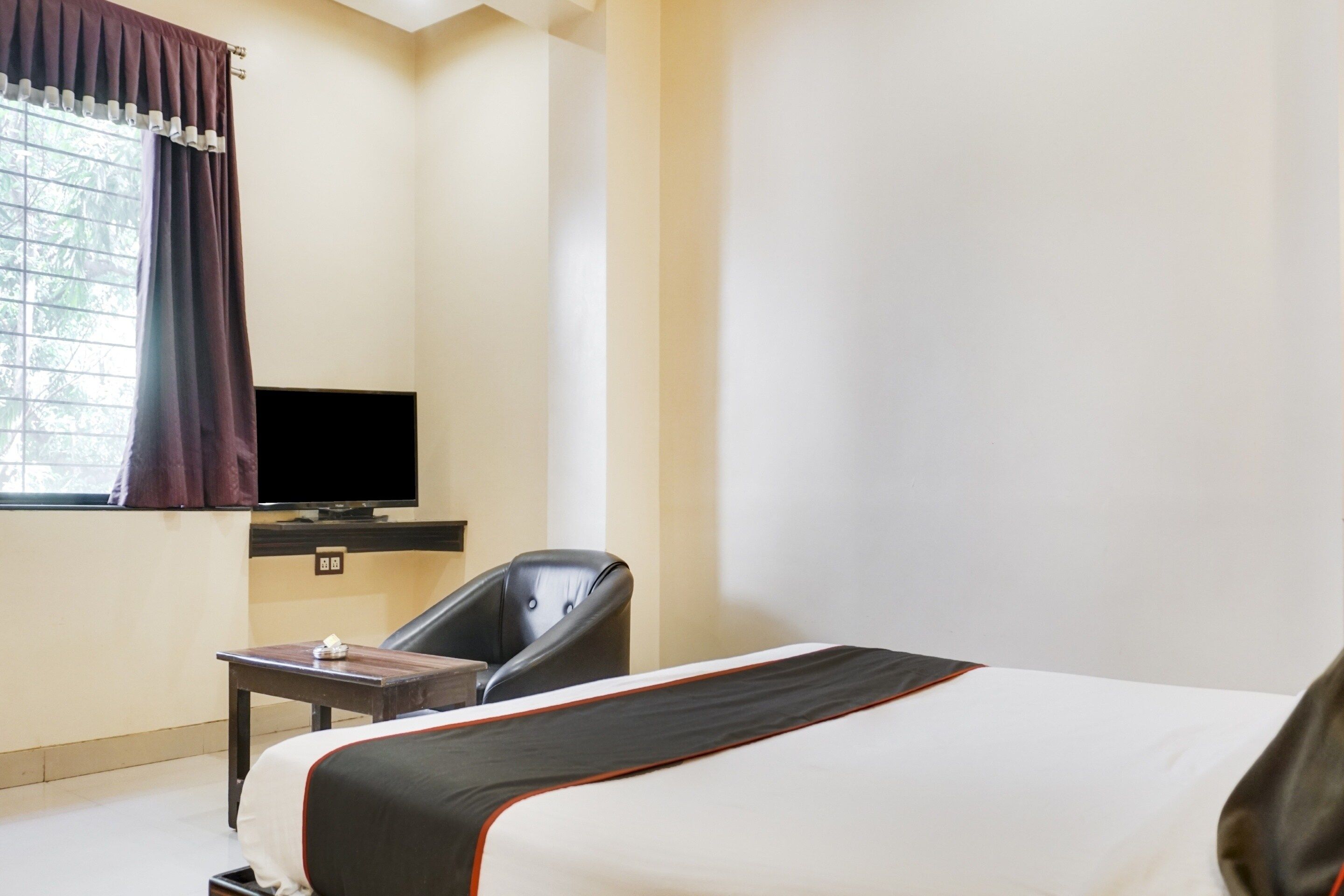 Quarto de hotel com cama, cadeira, mesinha, televisão e janela com cortinas.