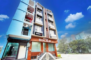 Um hotel de vários andares com um letreiro chamativo 'HOTEL BONJOUR', aparelhos de ar condicionado e uma escadaria que leva à entrada.