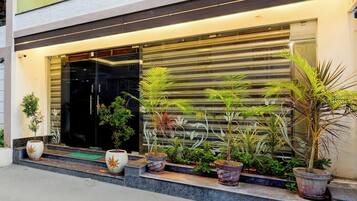 Entrada de hotel com design moderno, apresentando porta de vidro, portão de metal e diversos vasos com plantas.