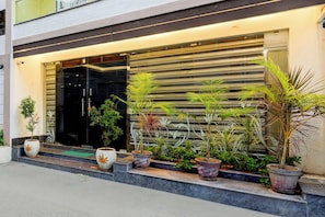 Entrada de hotel com design moderno, apresentando porta de vidro, portão de metal e diversos vasos com plantas.