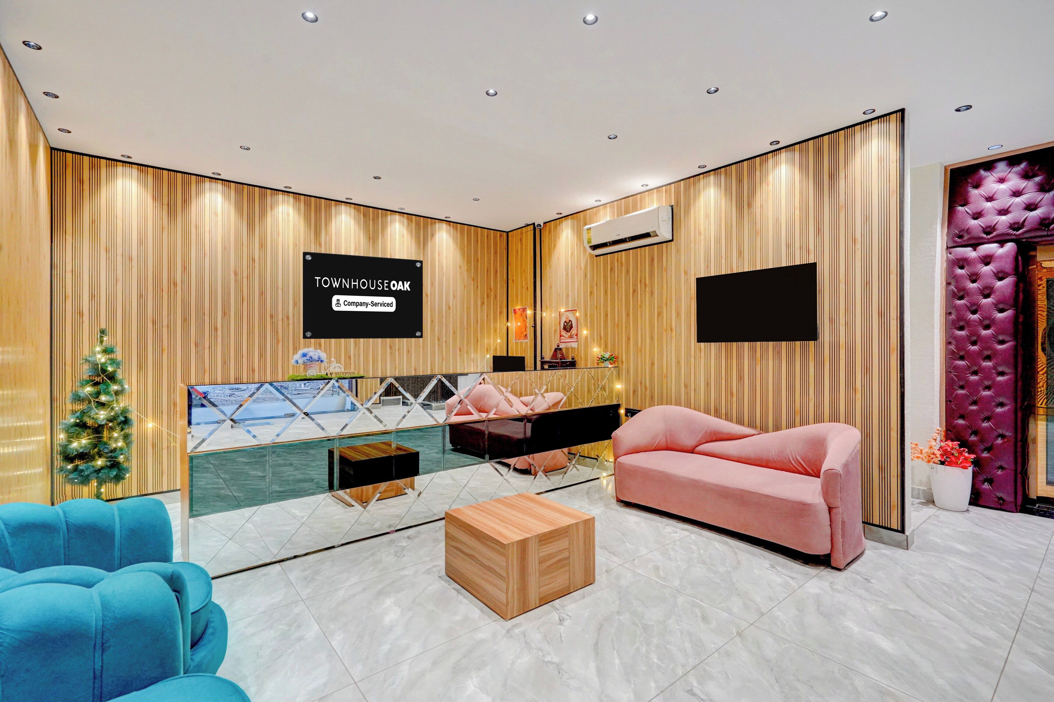 Uma área de recepção moderna com painel de madeira, televisão instalada, divisória de vidro e um espaço de estar com sofá rosa e poltrona azul.