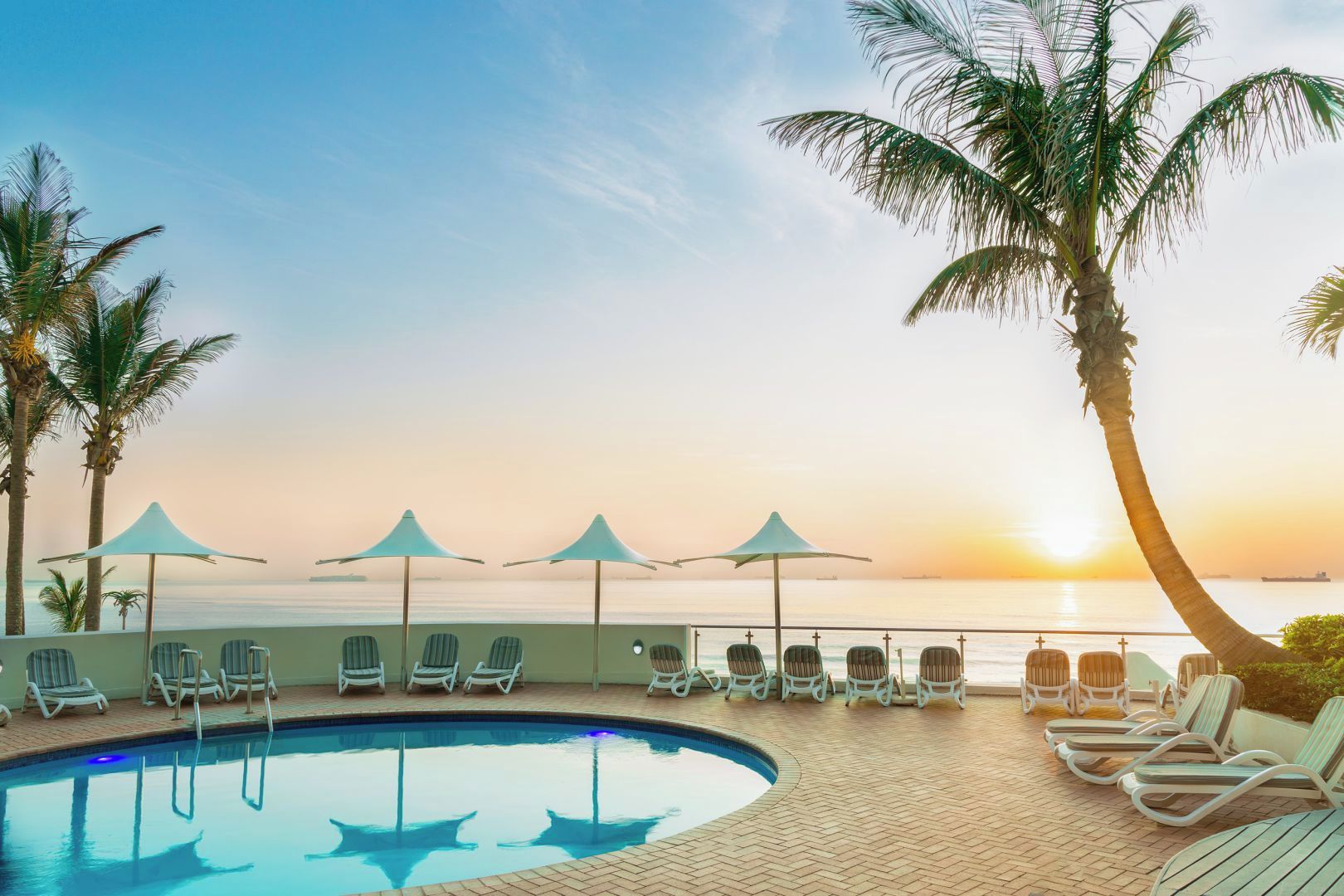 Área à beira da piscina com espreguiçadeiras e guarda-sóis, com vista para um pôr do sol sobre o mar.