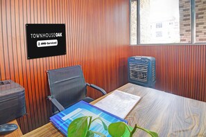 Quarto com parede de painel de madeira, uma placa preta com a inscrição 'TOWNHOUSE OAK OYO-Serviced', um ar condicionado azul, uma cadeira de escritório preta, uma mesa de madeira com uma planta e um caderno.
