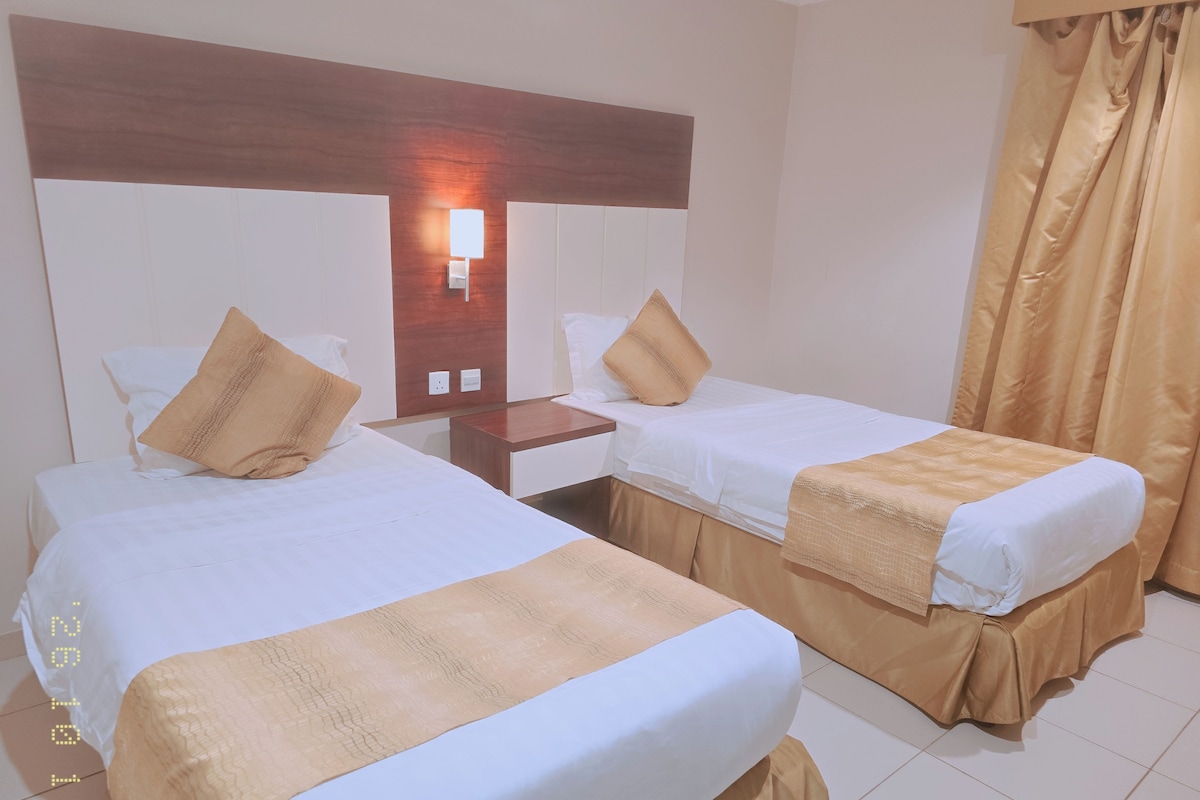 Quarto de hotel com duas camas, uma mesa de cabeceira, uma luminária na parede e uma cortina.